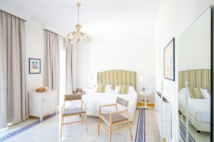 Quarto de hotel com uma cama grande, duas cadeiras, uma mesa de cabeceira e um espelho, além de um candelabro.