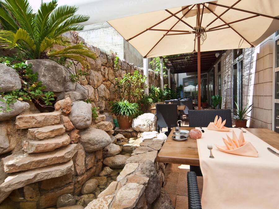 Um muro de pedra com plantas, uma mesa de madeira com toalha e um guarda-sol.