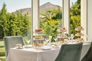 Uma mesa posta para um chá, com bandejas empilhadas de pâtisseries e sanduíches, copos de vidro elegantes e vista para uma montanha e vegetação.
