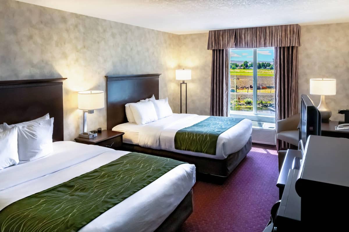 Quarto de hotel com duas camas, uma mesa, uma cadeira e uma janela grande com vista para o exterior.