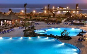 Uma área de piscina em resort, com várias piscinas, espreguiçadeiras, guarda-sóis e vista para o oceano.