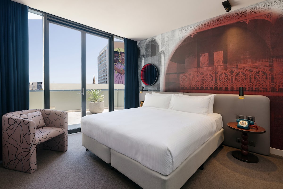 Quarto de hotel com uma cama grande, uma cadeira, uma mesinha com telefone e vista da cidade através de amplas janelas.