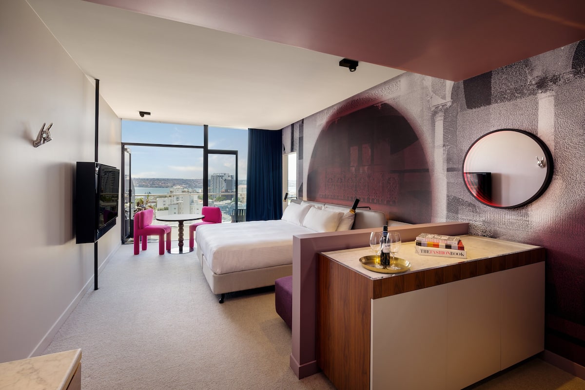 Quarto de hotel com cama, escrivaninha com espelho, televisão e vista para a cidade.