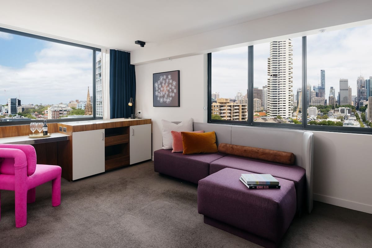 Quarto moderno com vista para a cidade, uma escrivaninha, uma cadeira rosa, um sofá roxo e um radiador.