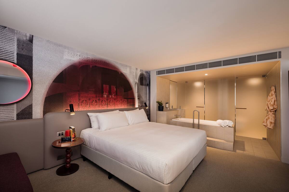 Quarto de hotel moderno com uma cama grande, uma pequena mesa de cabeceira com um relógio, uma luminária de mesa embutida, um espelho grande e um banheiro separado com banheira e chuveiro.