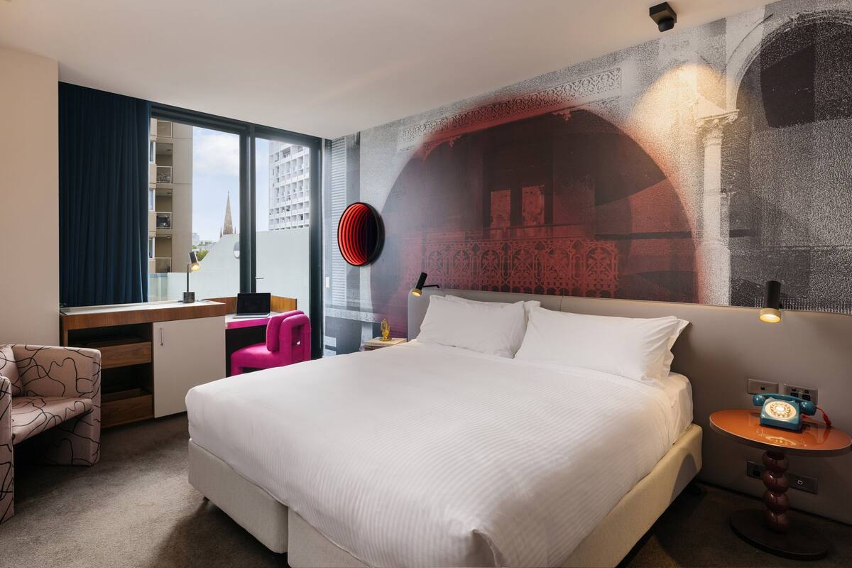 Quarto de hotel moderno com uma cama grande, uma escrivaninha, uma cadeira e vista para a paisagem urbana através de uma janela ampla.