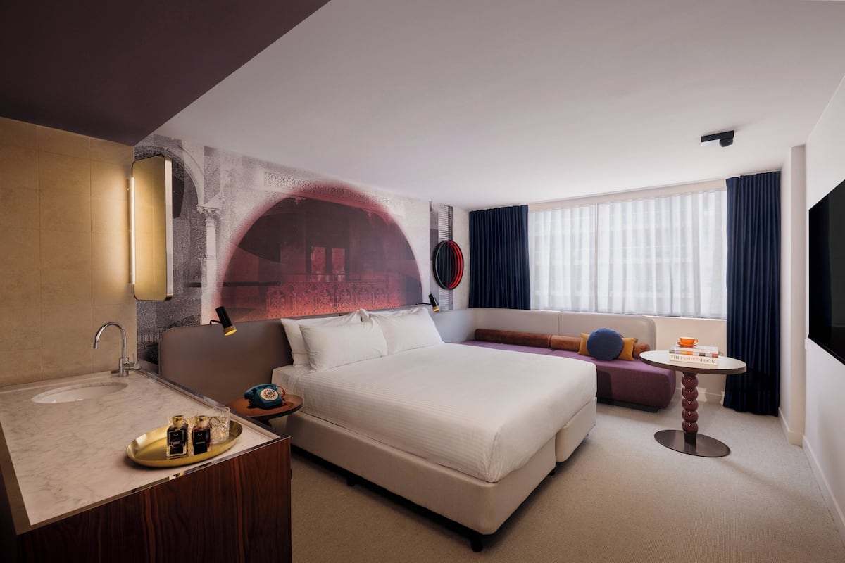 Quarto de hotel moderno com uma cama grande, um sofá pequeno, uma mesa redonda e um banheiro com pia e espelho.