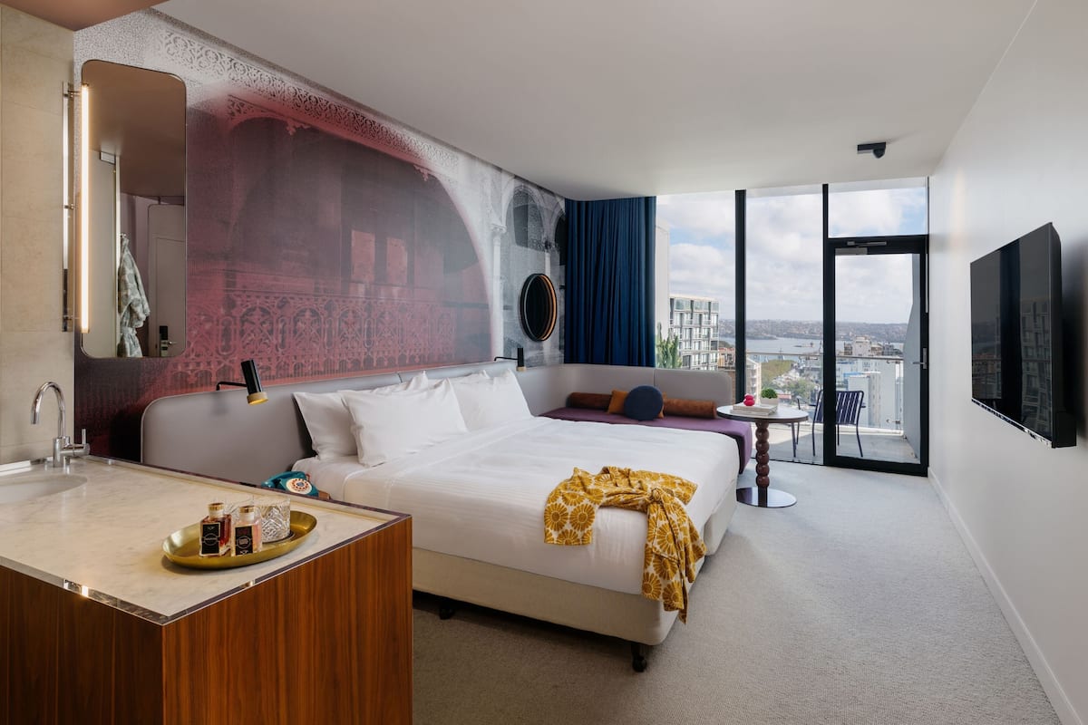 Quarto de hotel com uma cama grande, uma escrivaninha com espelho, uma mesa pequena e vista para a cidade através de grandes janelas.