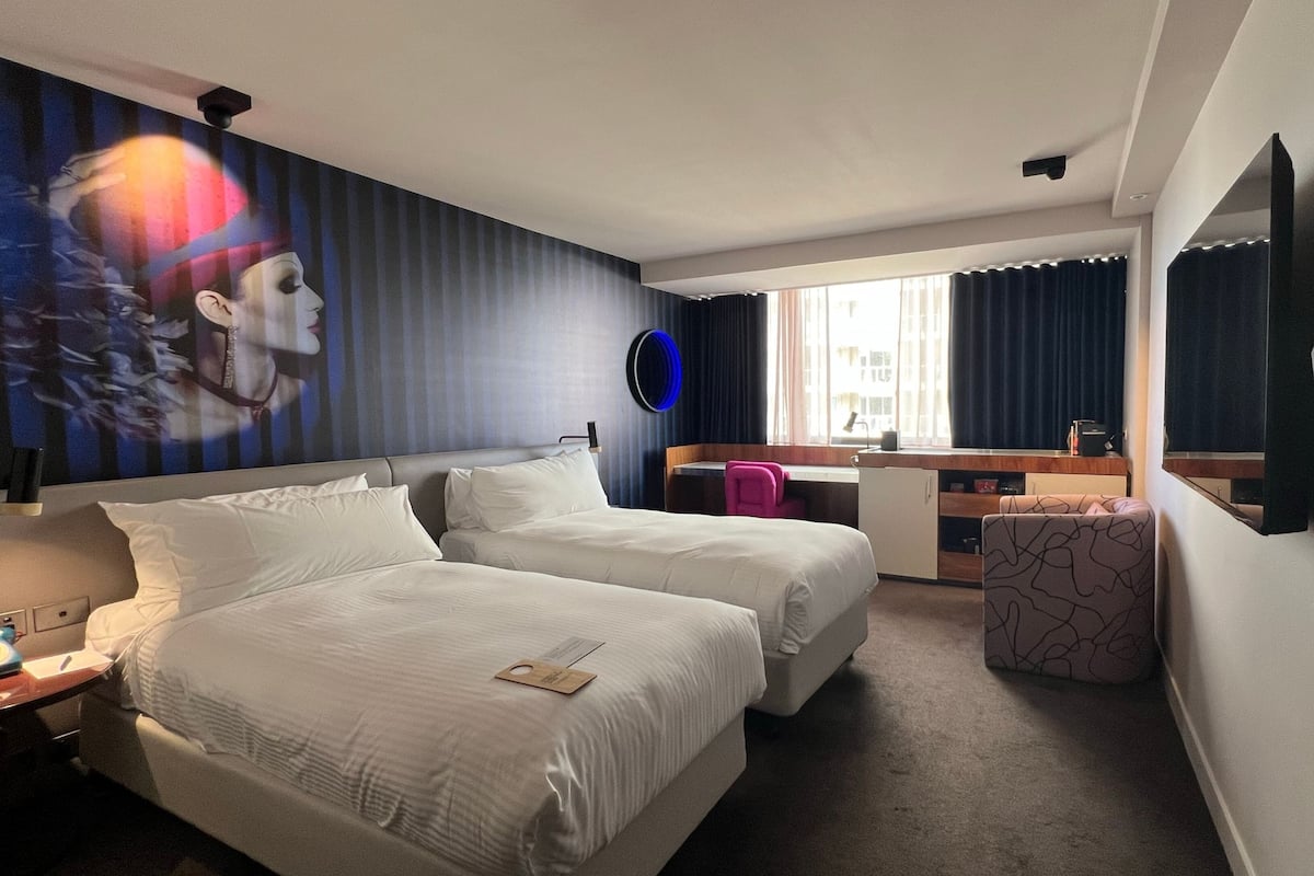 Quarto de hotel com duas camas, uma escrivaninha, uma cadeira, televisão e uma janela grande.