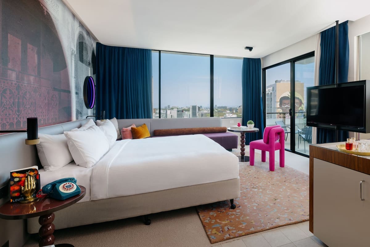 Quarto de hotel com uma cama grande, televisão, mesinha com telefone e vista da cidade através de janelas amplas.