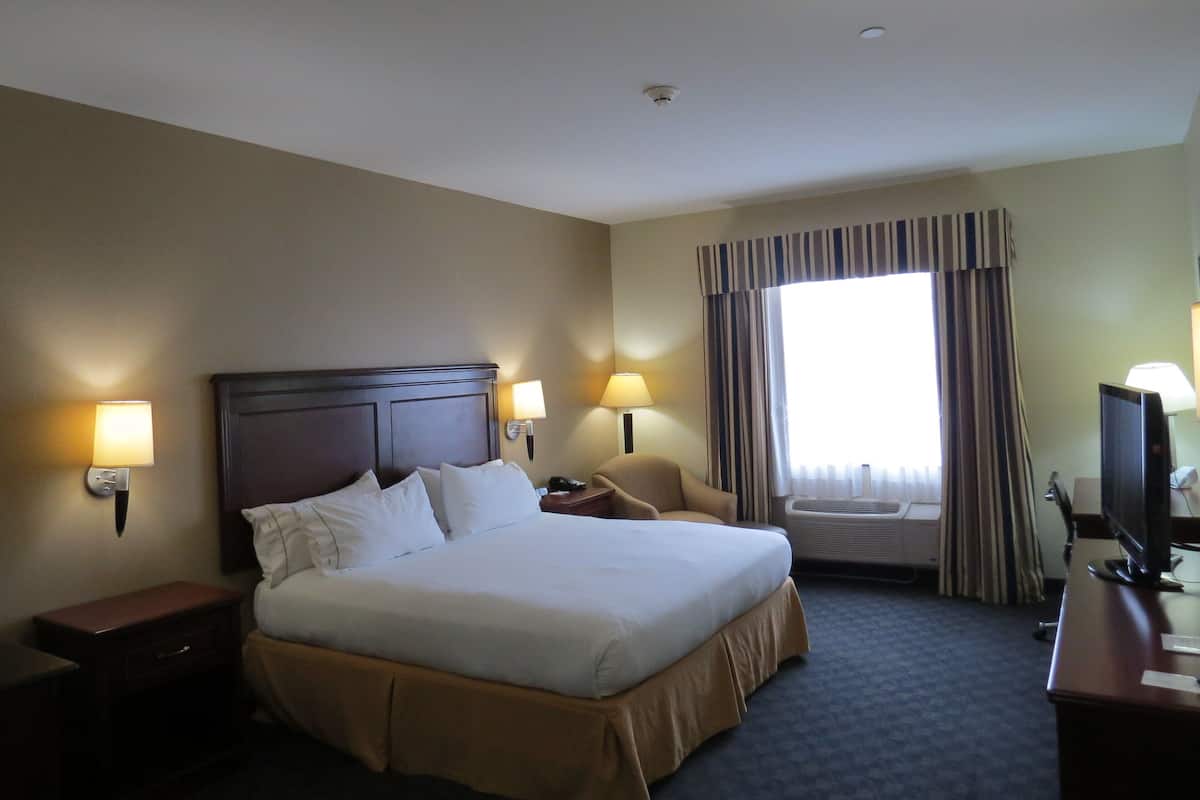 Quarto de hotel com uma cama grande, duas luminárias de cabeceira, uma mesa de trabalho com televisão e uma janela com cortinas.