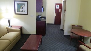 Quarto de hotel com sofá, uma pequena mesa, escrivaninha com computador, televisão e porta para um banheiro.
