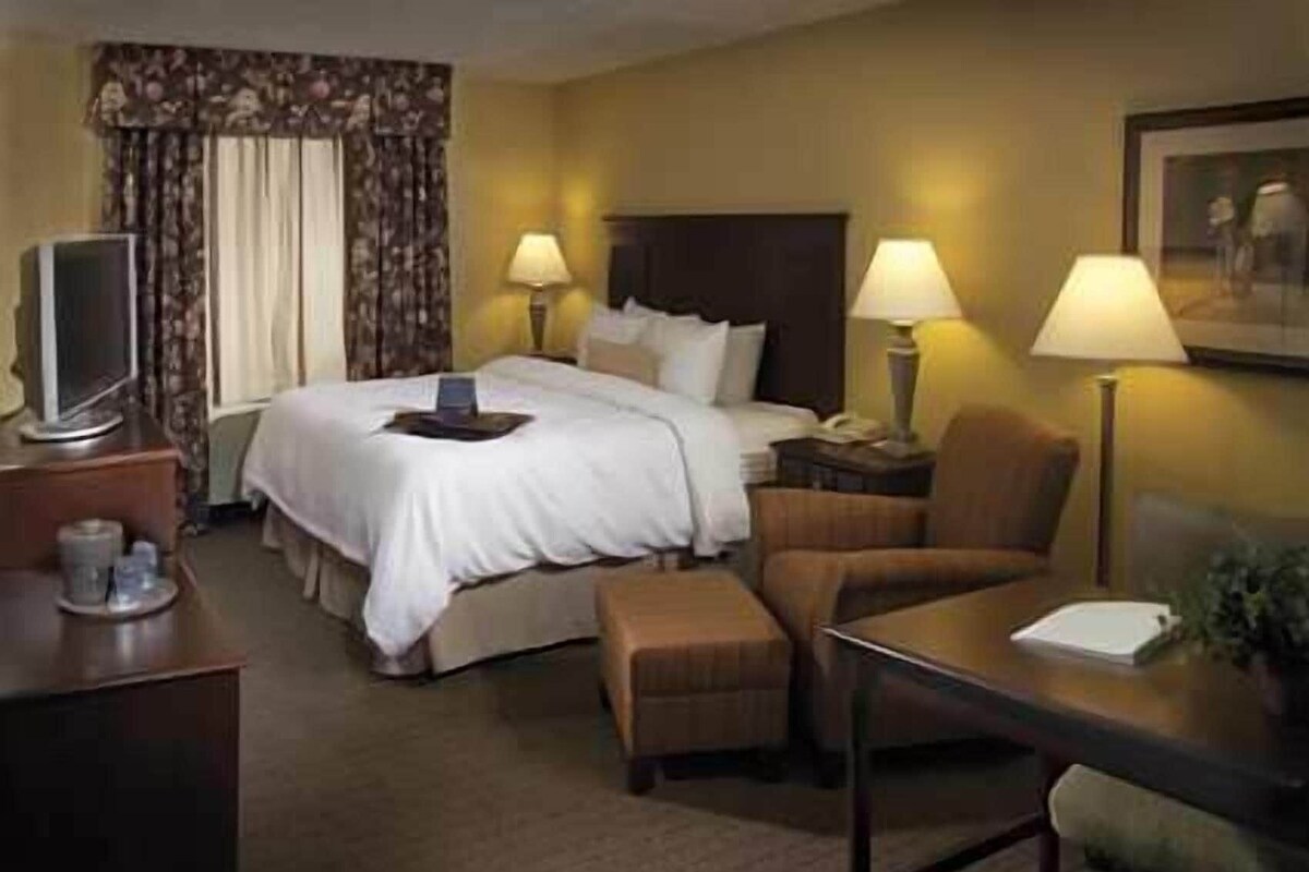 Quarto de hotel com uma cama, duas luminárias de cabeceira, uma televisão, uma escrivaninha e uma cadeira.