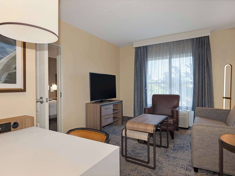 Quarto de hotel com televisão de tela plana, sofá, poltrona, mesa pequena e luminária.