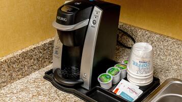 Máquina de café Keurig com bandeja coletora, cápsulas de café e uma xícara reutilizável sobre uma bancada de granito.