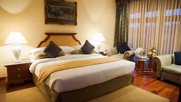 Quarto de hotel com uma cama grande, duas mesas de cabeceira com luminárias, uma cadeira, uma pequena mesa com um vaso de flores e um quadro na parede.