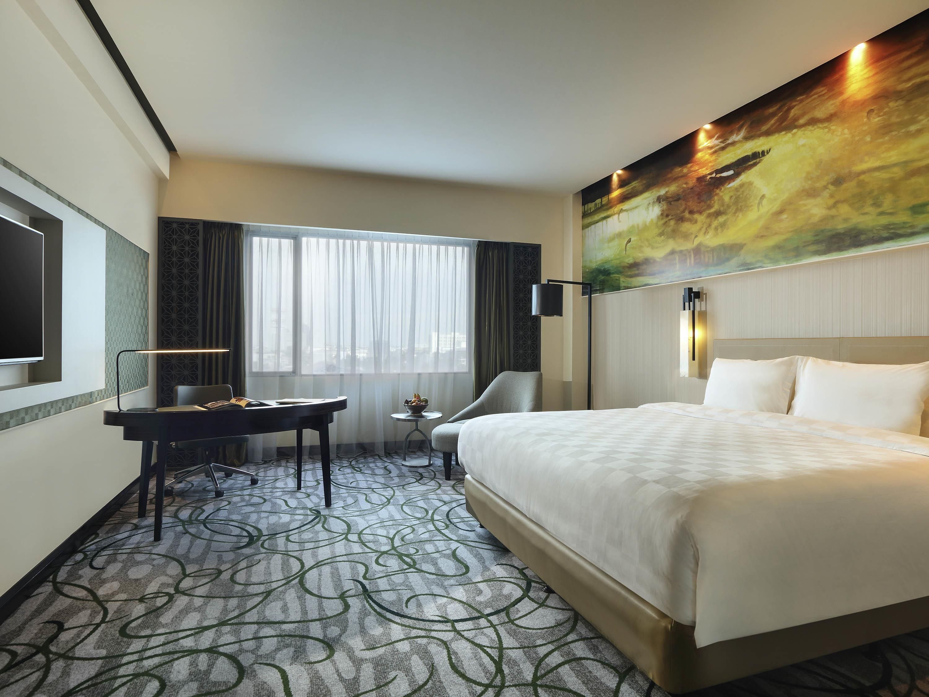 Quarto de hotel com cama, escrivaninha com cadeira, mesinha de cabeceira com abajur e uma grande janela com cortinas.