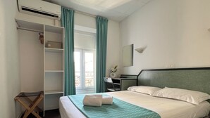 Quarto de hotel com cama, escrivaninha, cadeira, guarda-roupa e janela com cortinas.