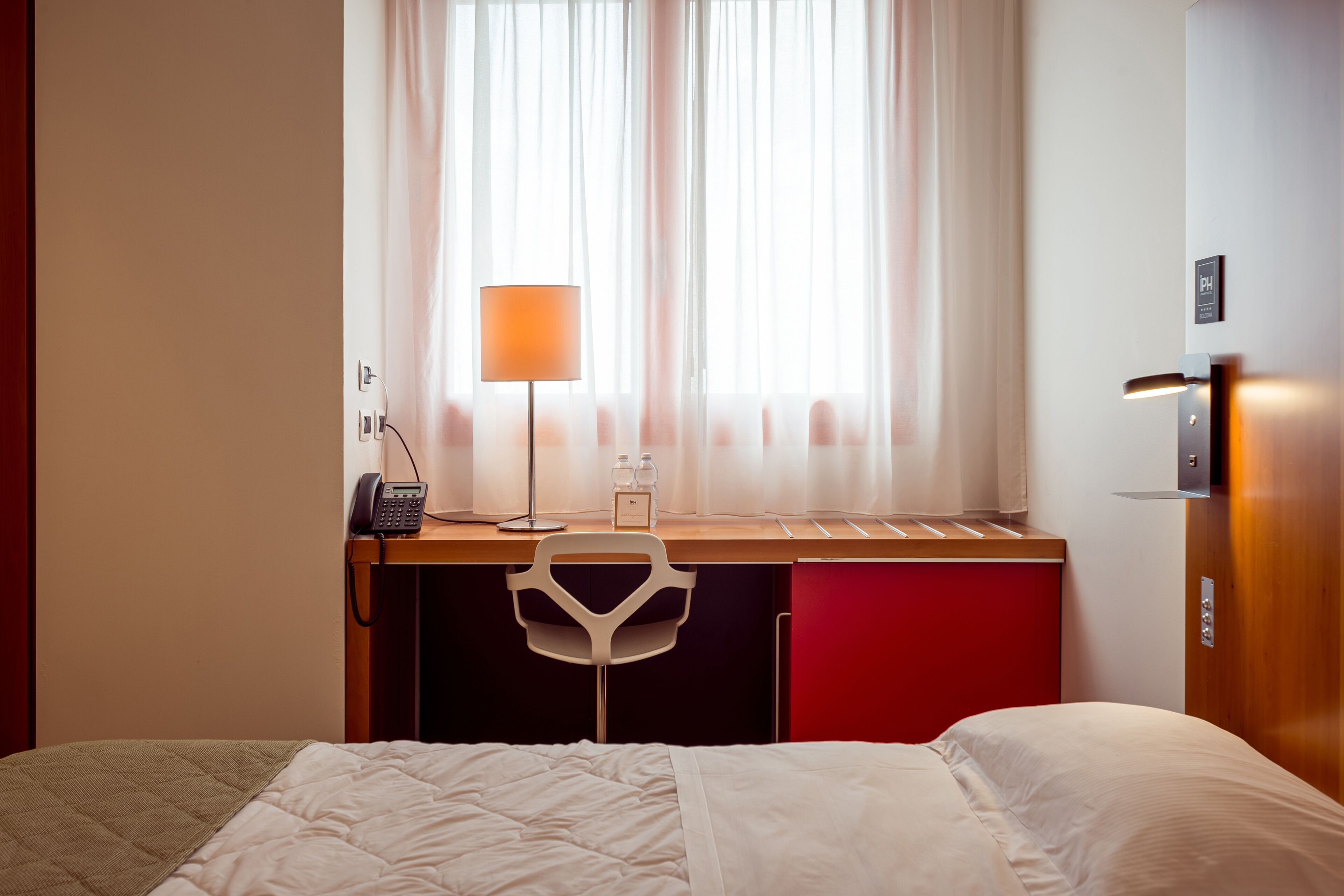 Quarto de hotel com cama, escrivaninha com luminária, cadeira e janela com cortinas.