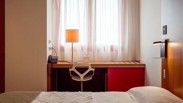 Quarto de hotel com cama, escrivaninha com luminária, cadeira e janela com cortinas.