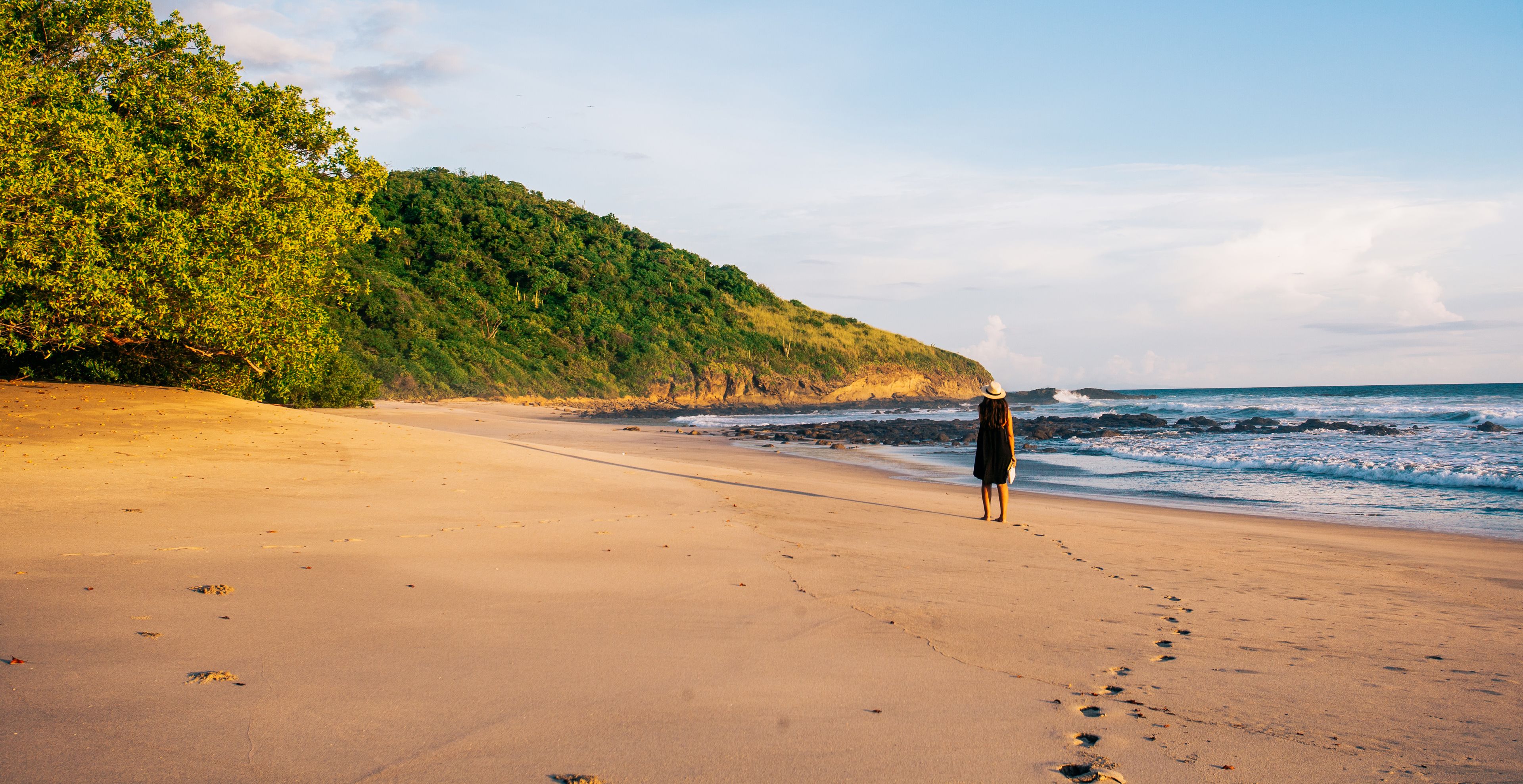 Uma praia com uma pessoa caminhando pela margem, areia dourada e um morro coberto de vegetação.