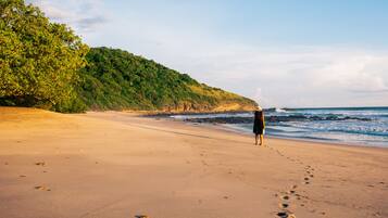 Uma praia com uma pessoa caminhando pela margem, areia dourada e um morro coberto de vegetação.