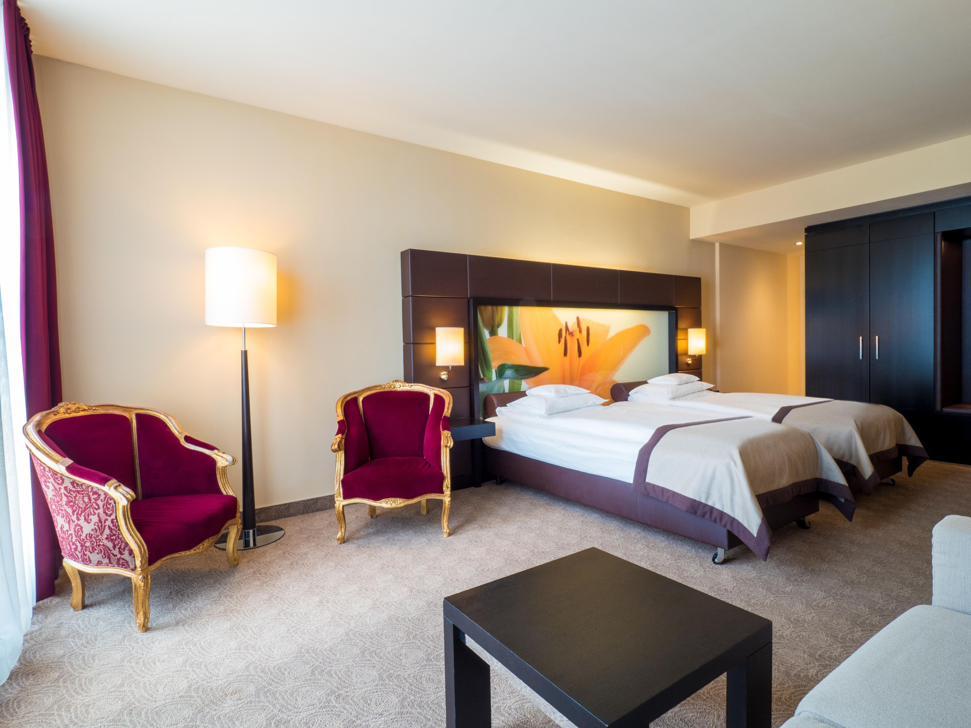 Quarto de hotel com uma cama grande, duas poltronas, uma mesinha e cabeceira com padrão floral.