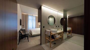 Quarto de hotel moderno com uma cama grande, uma mesinha de cabeceira, uma escrivaninha, uma cadeira, um espelho redondo, uma pia e um guarda-roupa.