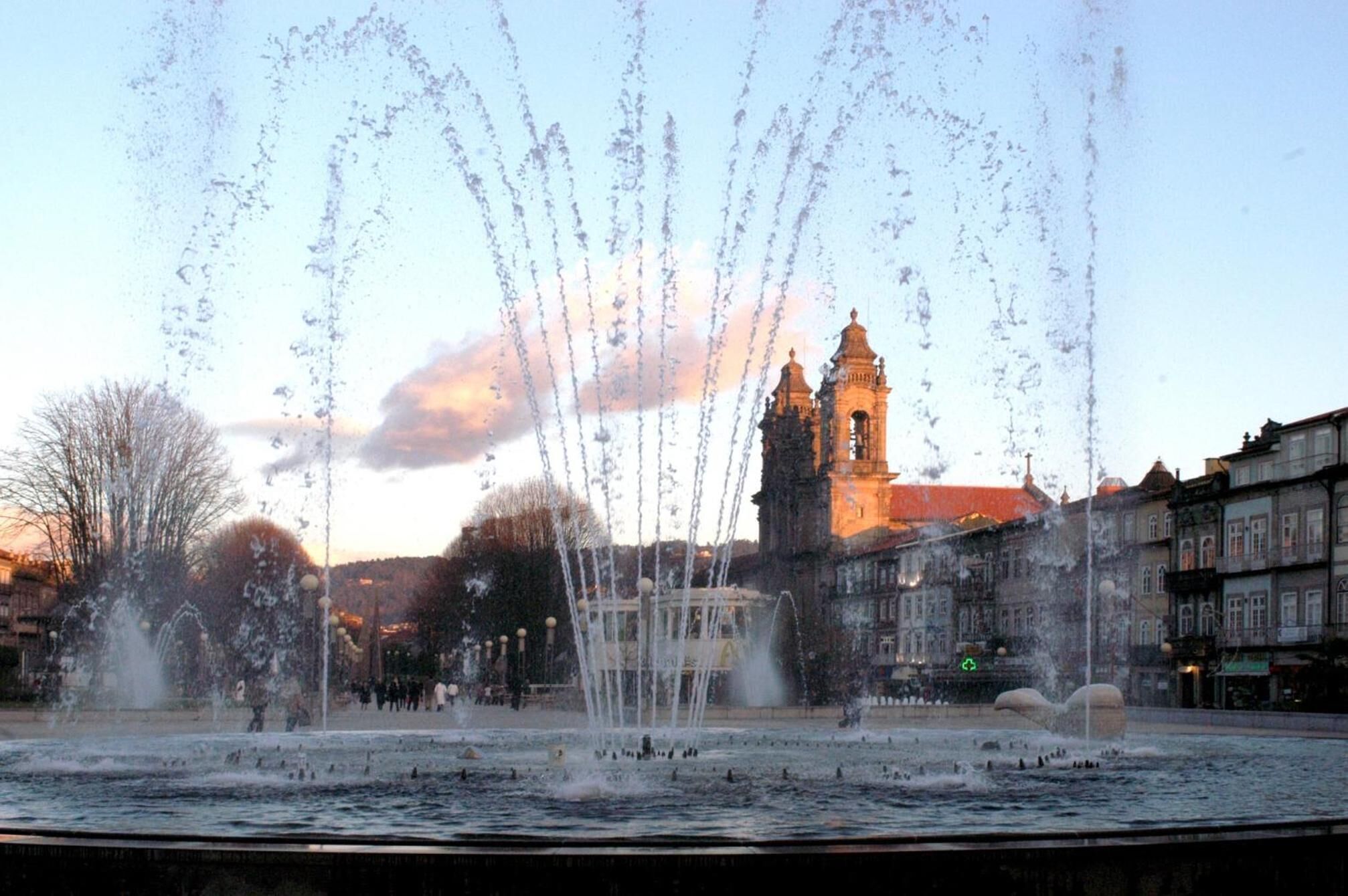 Uma fonte com vários jatos de água numa praça pública, com um edifício histórico ao fundo.