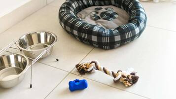 Uma cama para animais de estimação, comedouros de metal e um brinquedo em piso de azulejos.