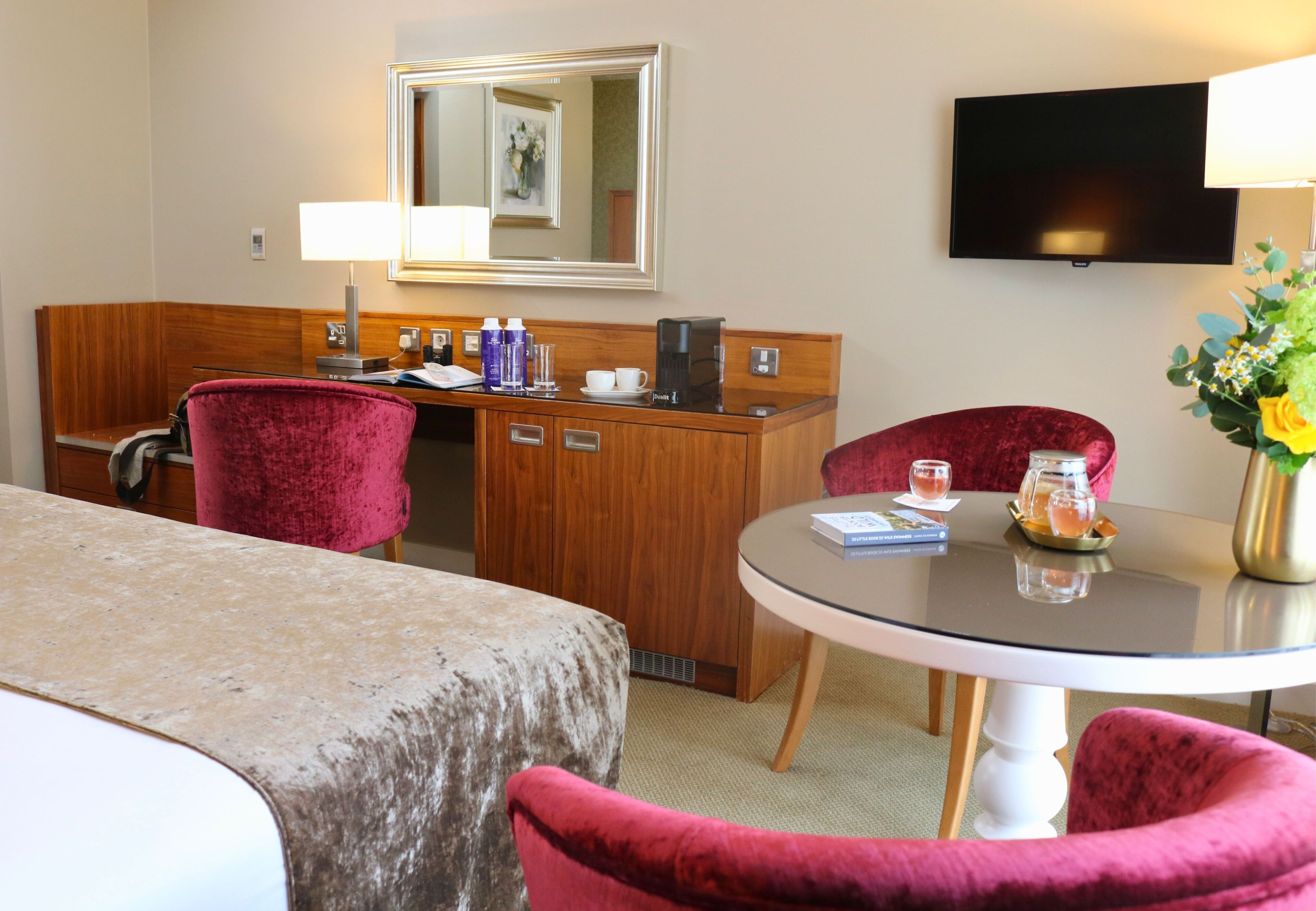 Quarto de hotel com cama, escrivaninha com espelho, televisão, mesa redonda com vaso e duas cadeiras.