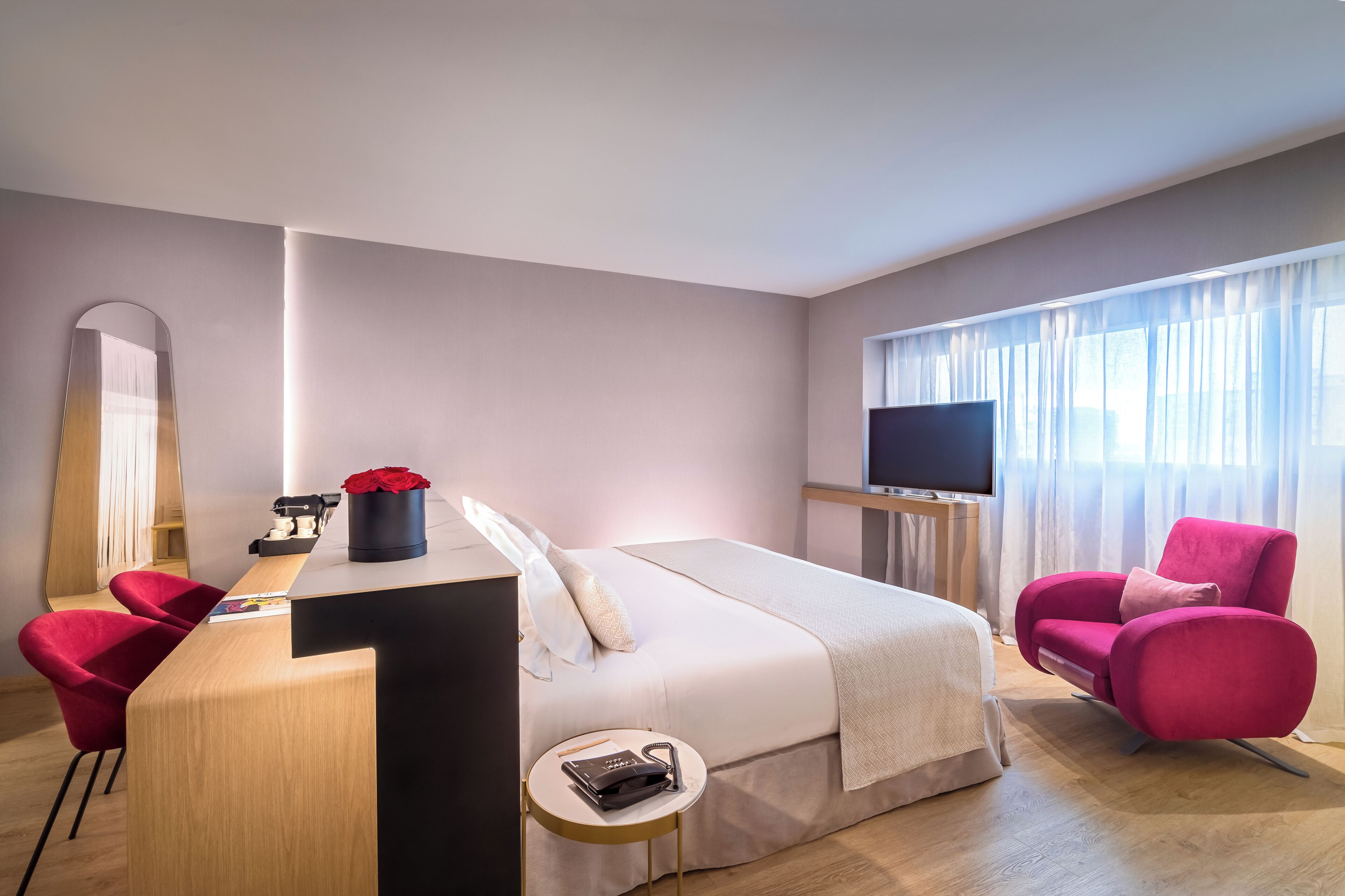 Quarto de hotel moderno com uma cama, escrivaninha, cadeira, televisão e uma janela ampla com cortinas.