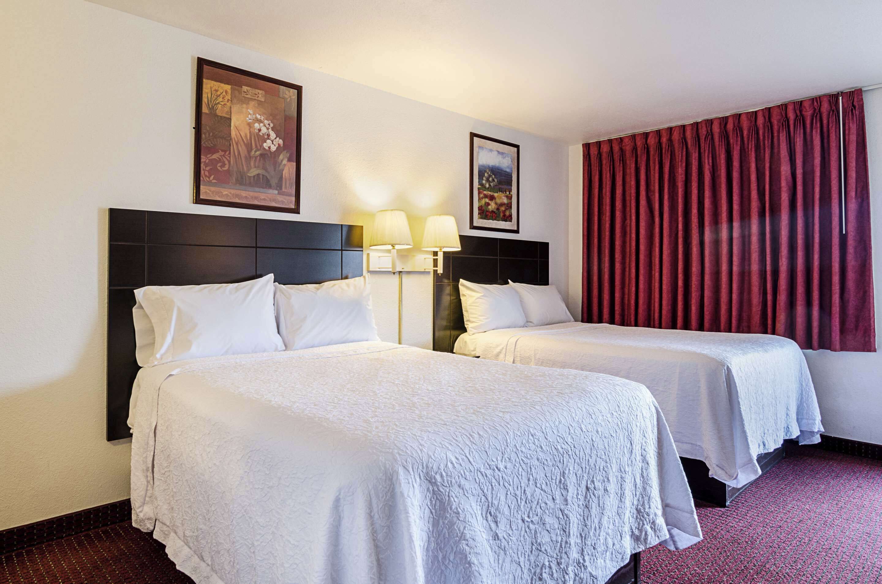Quarto de hotel com duas camas, cabeceira, luminárias fixadas na parede e uma janela grande com cortinas vermelhas.