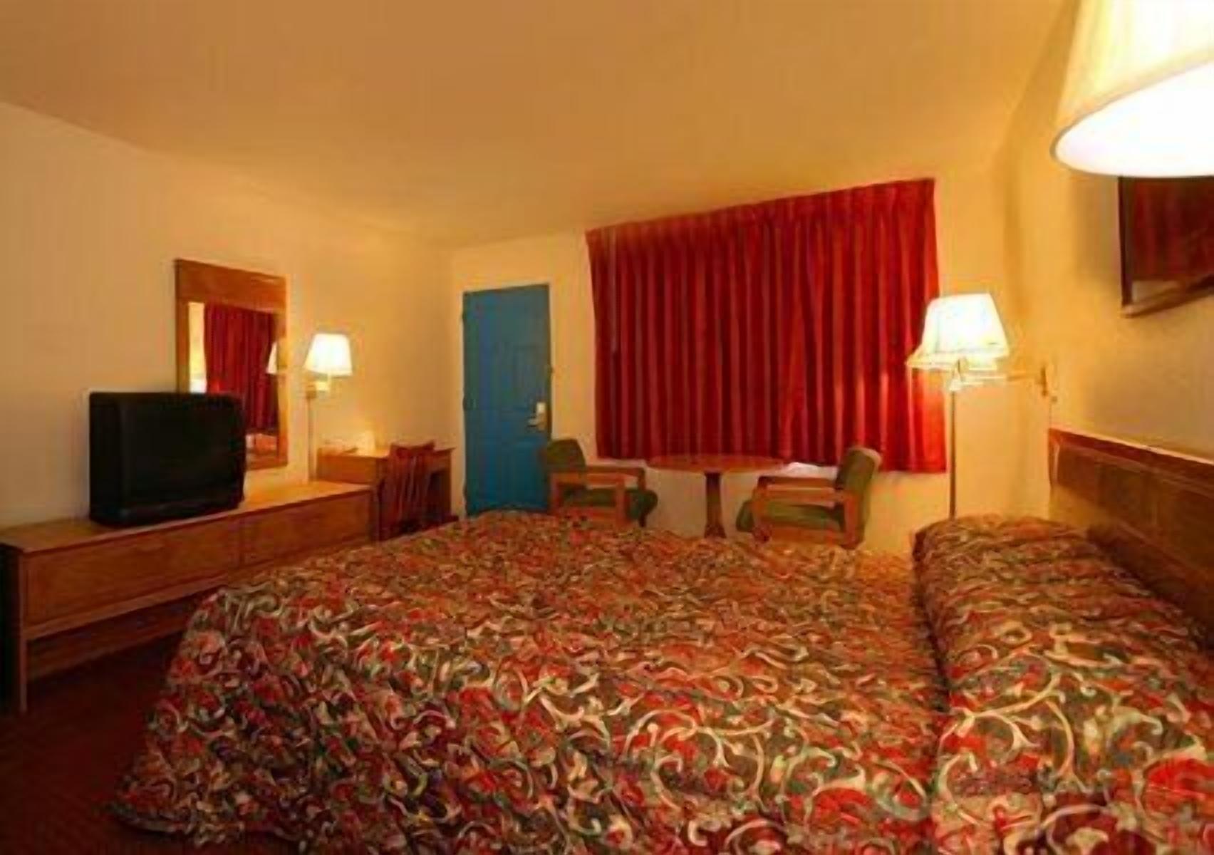 Quarto de hotel com uma cama grande, uma televisão em uma cômoda de madeira, uma escrivaninha com cadeira e uma janela com cortinas vermelhas.