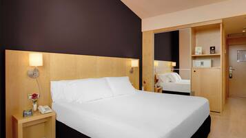 Quarto de hotel com uma cama grande, abajures, mesa de cabeceira com telefone e um vaso de flores, espelho e armário de madeira com micro-ondas.