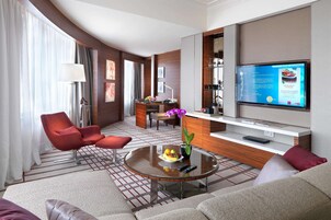 Quarto de hotel moderno com sofá, poltrona e mesa de centro redonda com frutas. Uma TV de tela plana está montada na parede, e há uma pequena escrivaninha com abajur.
