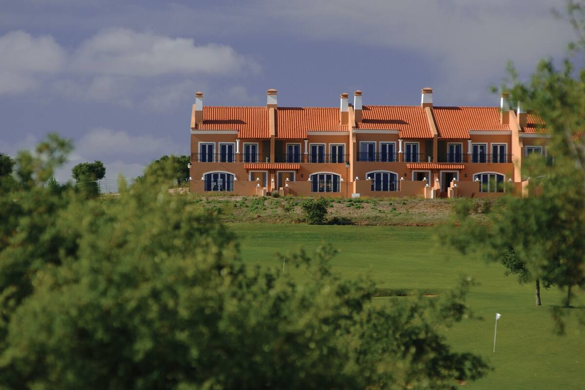 Um edifício grande, com vários andares, telhado de telhas vermelhas e diversas janelas, rodeado por vegetação e um campo de golfe.