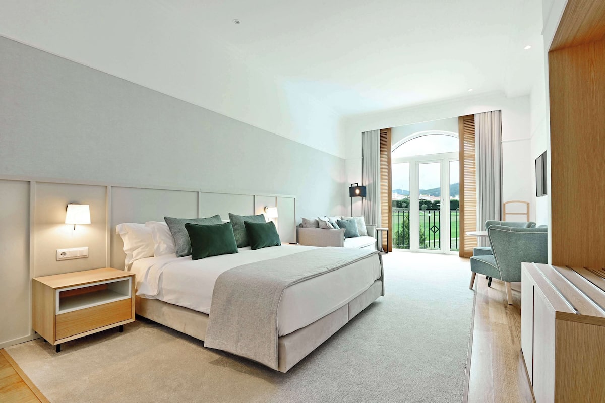Quarto de hotel moderno com cama grande, mesa de cabeceira, área de estar com uma poltrona e vista para o exterior.