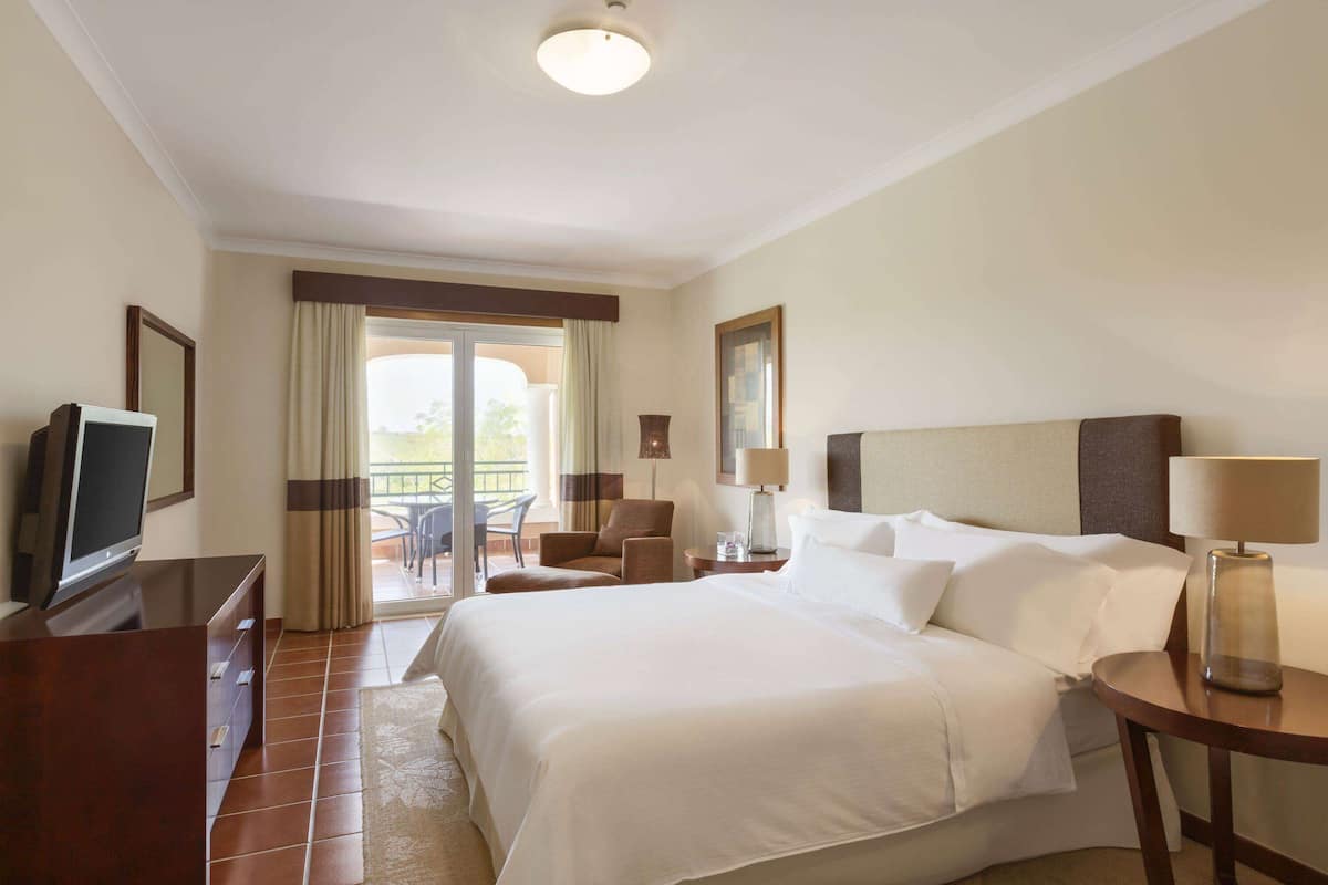 Quarto de hotel com uma cama grande, televisão, escrivaninha, cadeira, abajur e varanda com mesa e cadeiras.