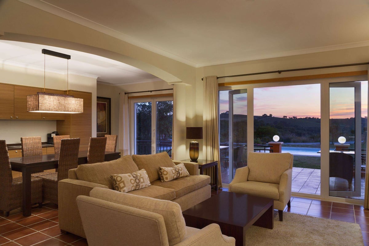 Uma sala de estar ampla com sofá, poltronas e uma mesa de jantar. Há uma janela grande com vista para o exterior.
