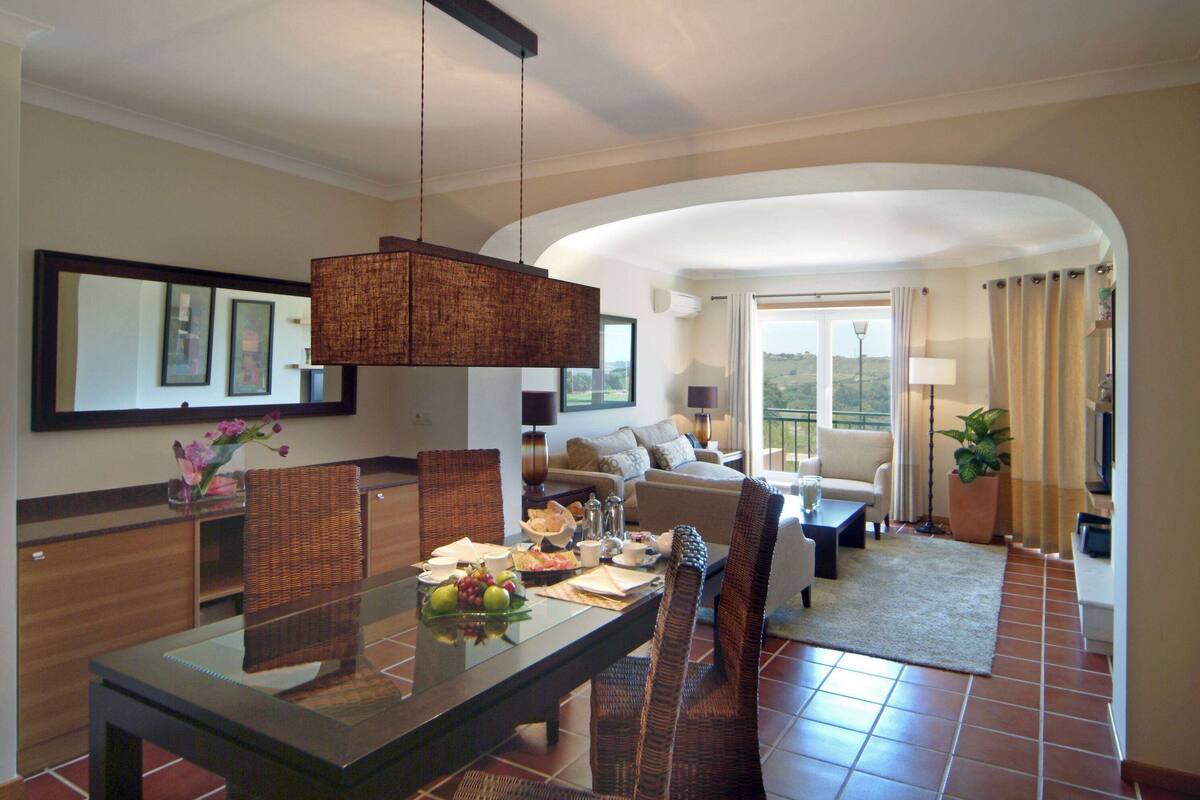 Uma sala de jantar com mesa de vidro, cadeiras de vime e luminária pendente.