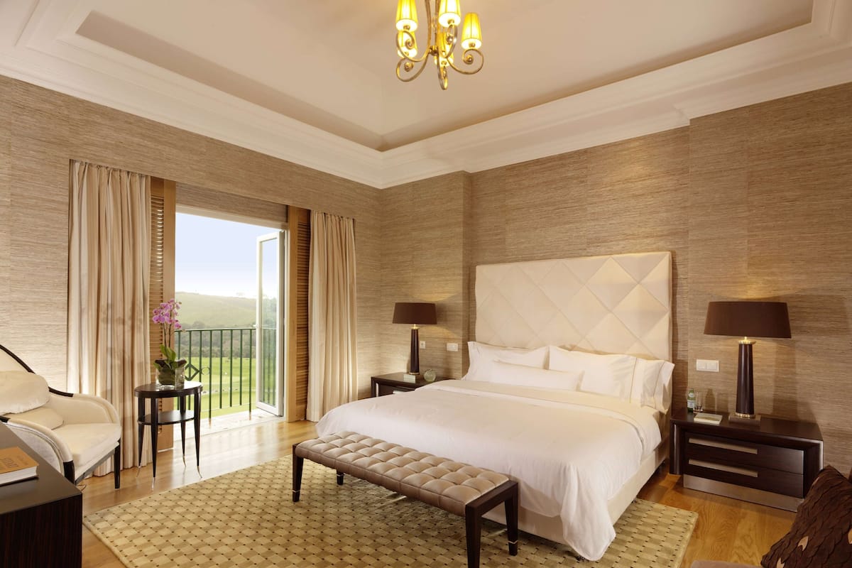 Um quarto espaçoso com uma cama grande, duas mesas de cabeceira com luminárias, um banquinho, uma pequena mesa com um vaso e vista para o exterior através de uma grande janela.
