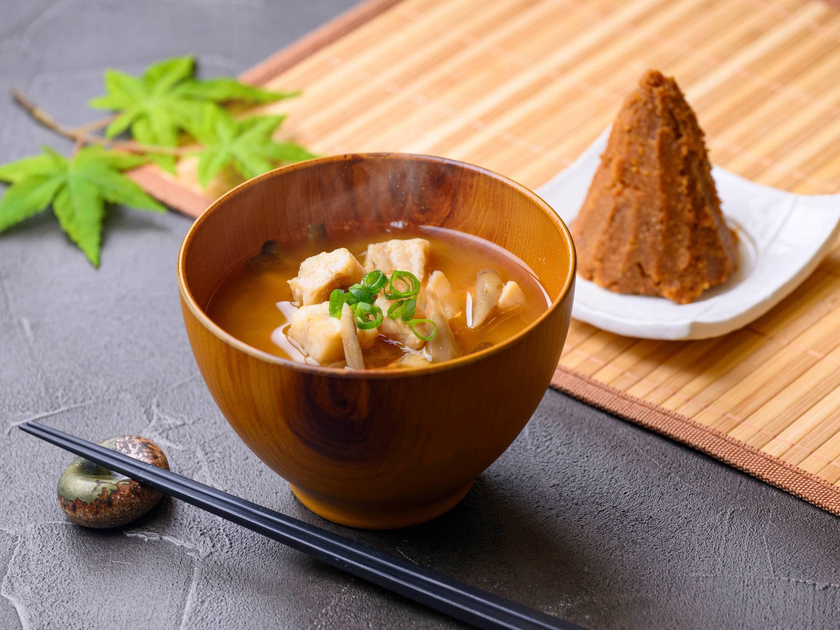 Uma tigela de sopa com tofu e cebolinha acompanhada por um cone que parece ser uma sobremesa, sobre uma superfície texturizada, com hashis e uma pedra.