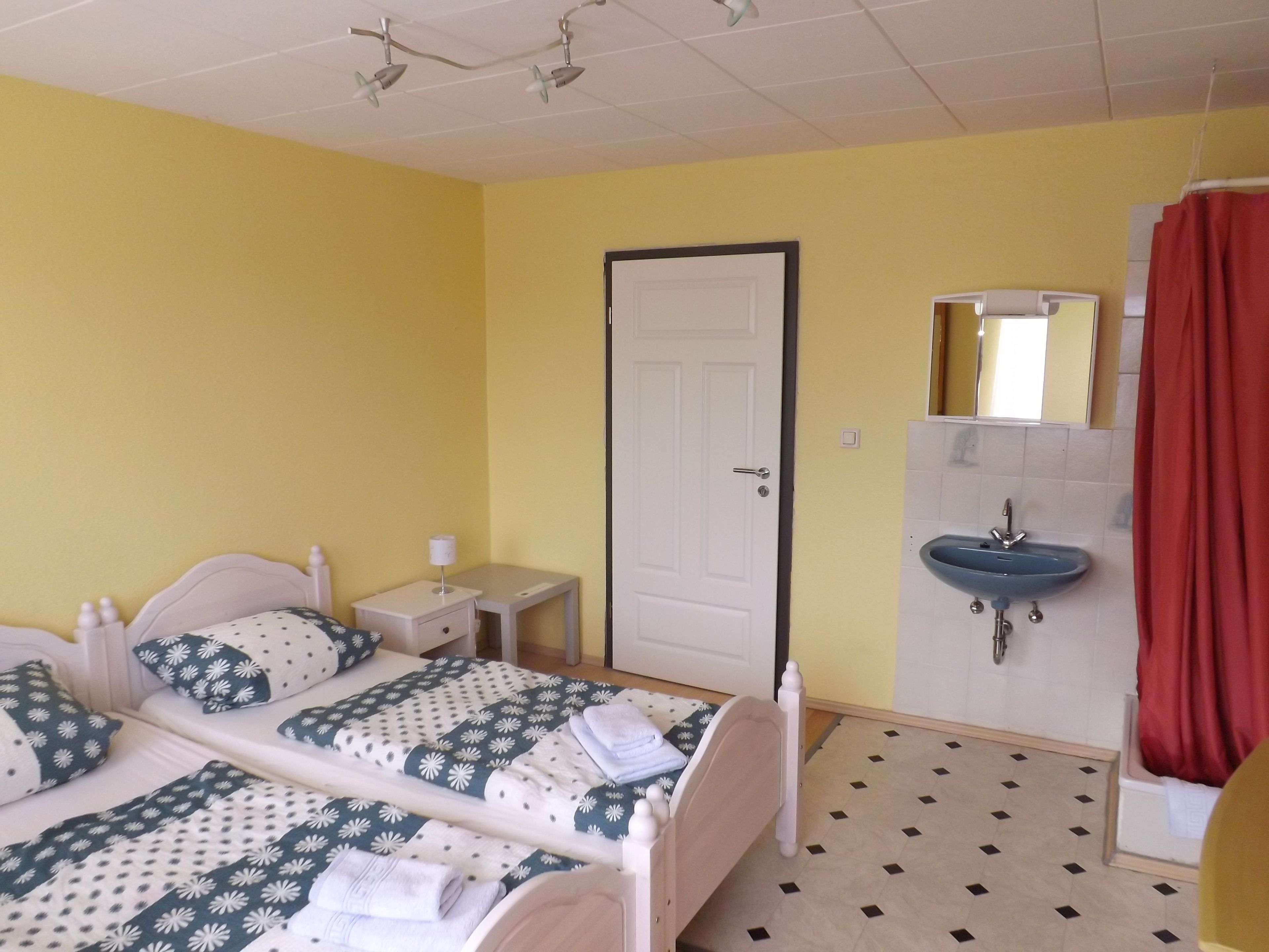 Quarto com duas camas, uma mesa de cabeceira, um lavatório e espelho na casa de banho, e uma cortina vermelha.
