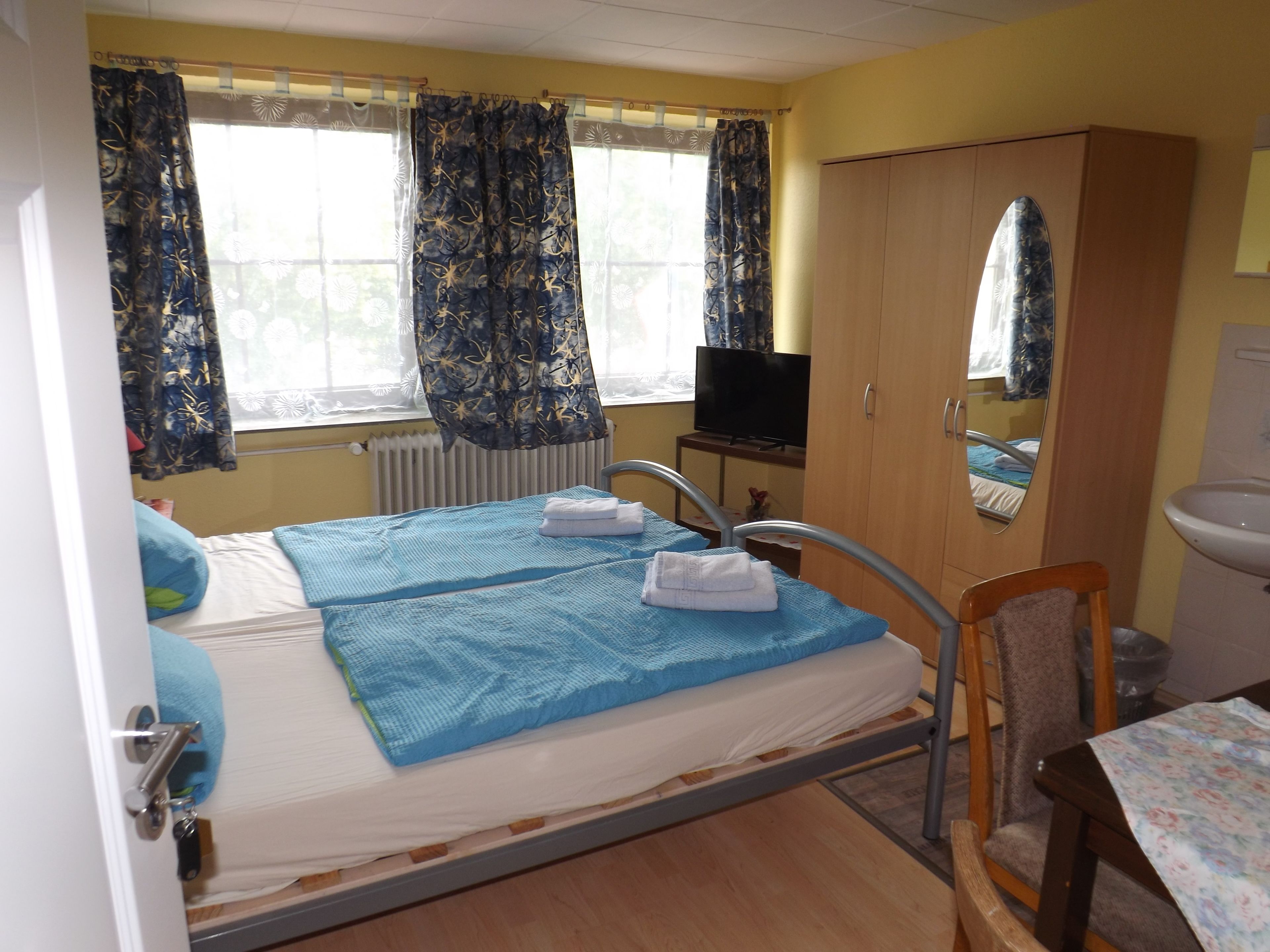 Um quarto de hospital com uma cama, uma televisão, um guarda-roupa de madeira, uma cadeira e uma pia.