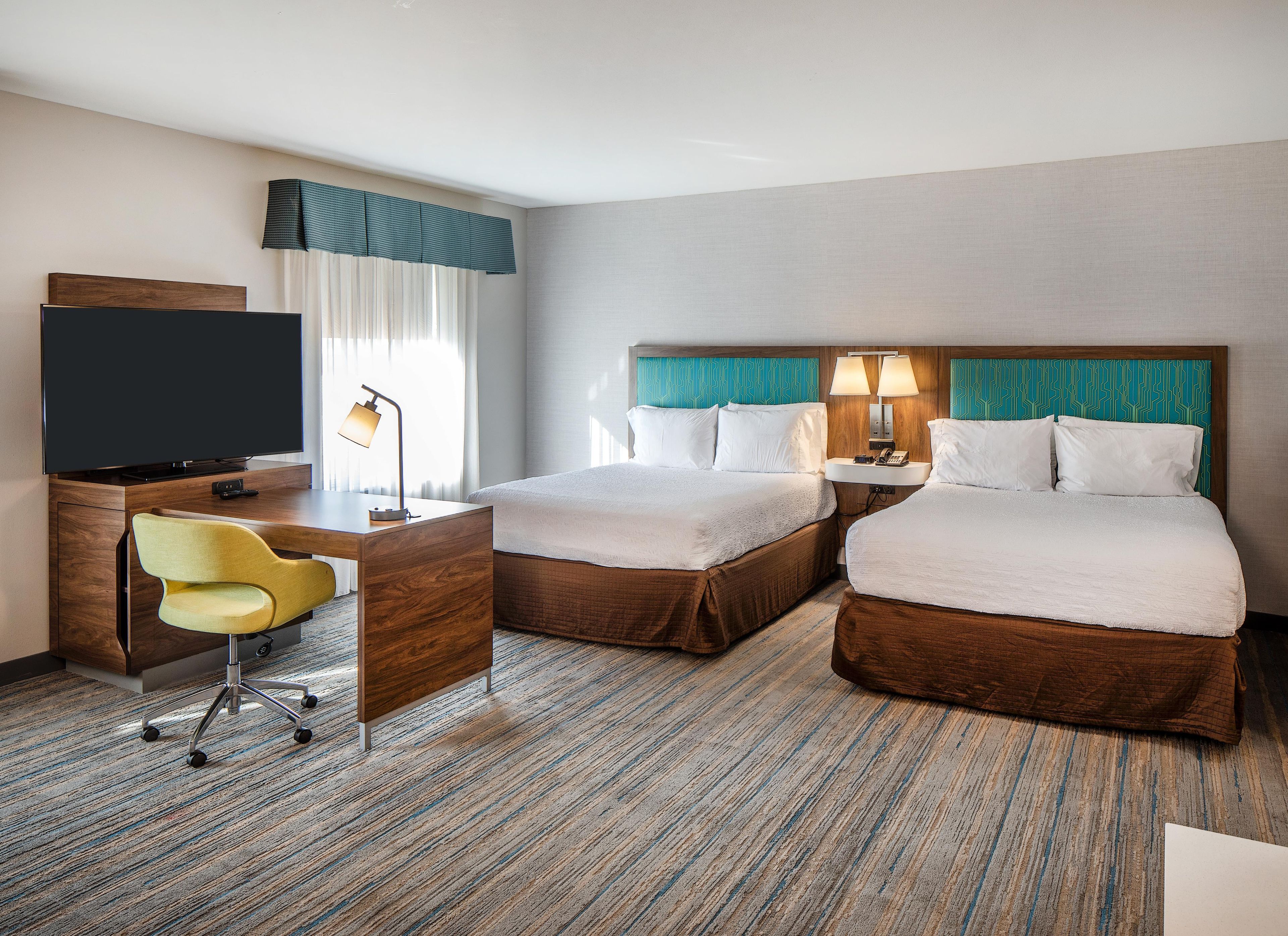 Quarto de hotel com duas camas, uma escrivaninha com televisão, uma cadeira e uma mesa de cabeceira com luminárias.