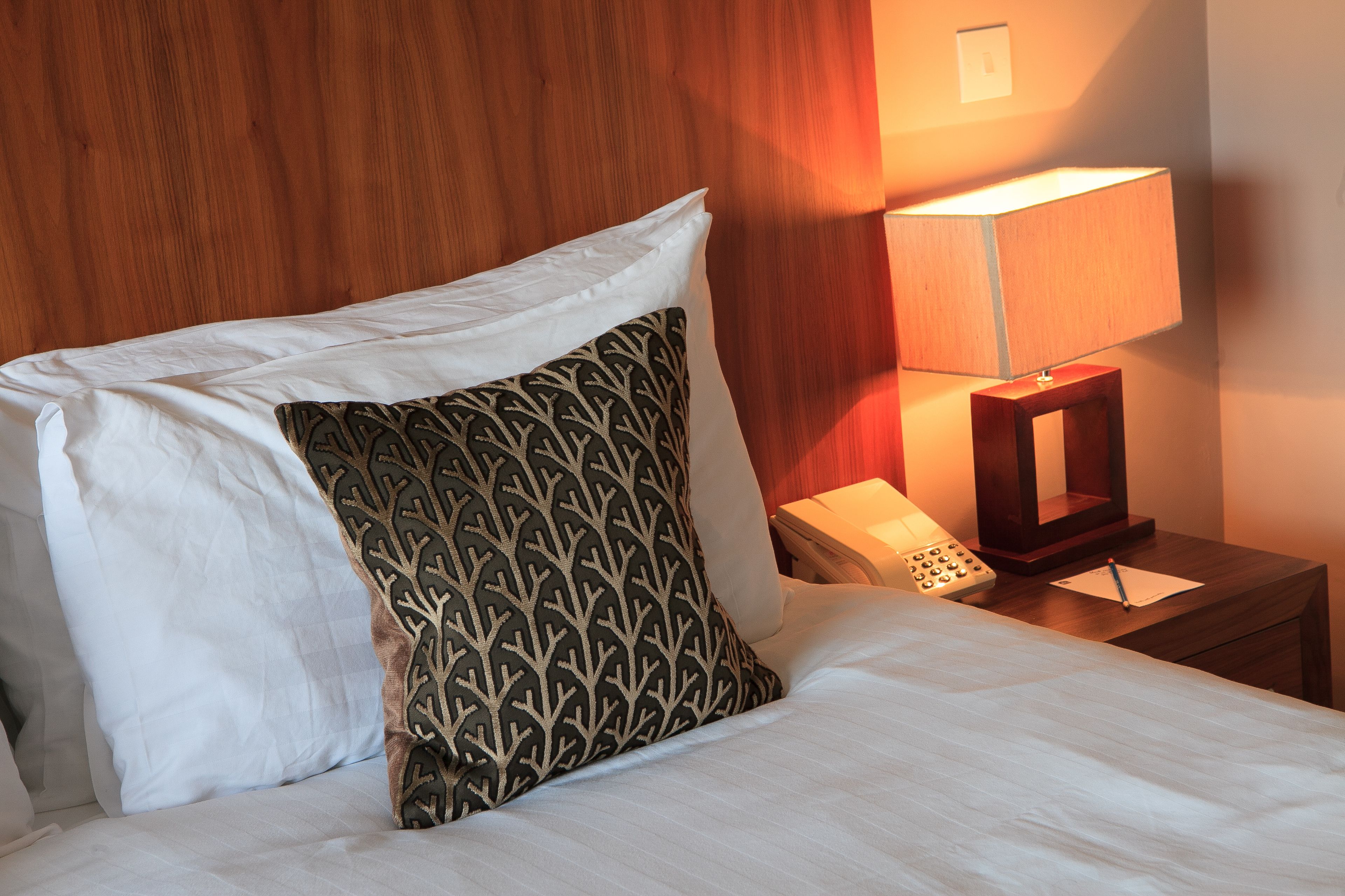 Cama bem arrumada com almofadas brancas e estampadas, um abajur e um telefone num criado-mudo de madeira.