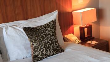 Cama bem arrumada com almofadas brancas e estampadas, um abajur e um telefone num criado-mudo de madeira.
