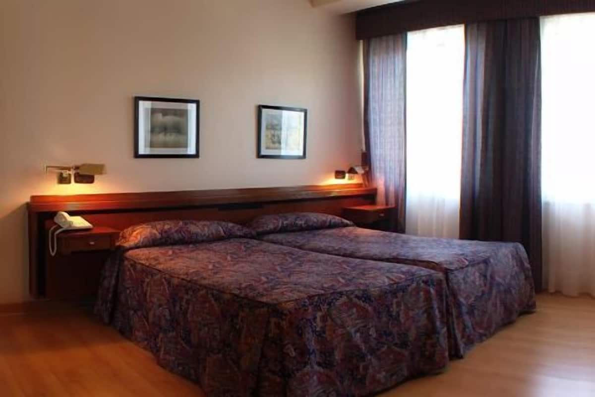 Uma cama de casal com colcha estampada, uma mesa de cabeceira com abajur e duas molduras na parede.