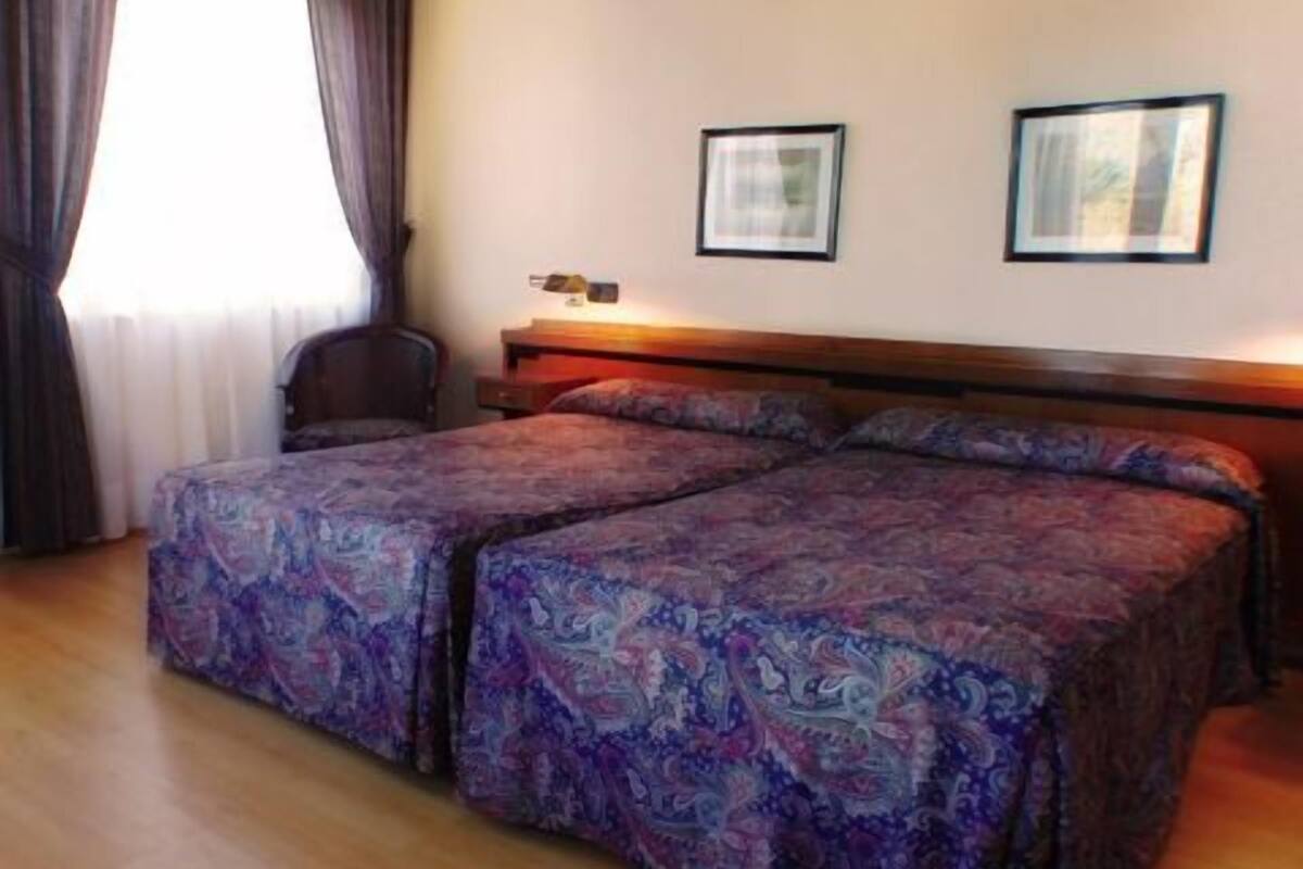 Quarto duplo com duas camas, cabeceira de madeira, uma cadeira e duas imagens emolduradas na parede.